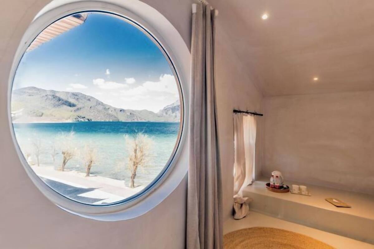 Quarto com janela redonda que oferece vista para um lago e montanhas.