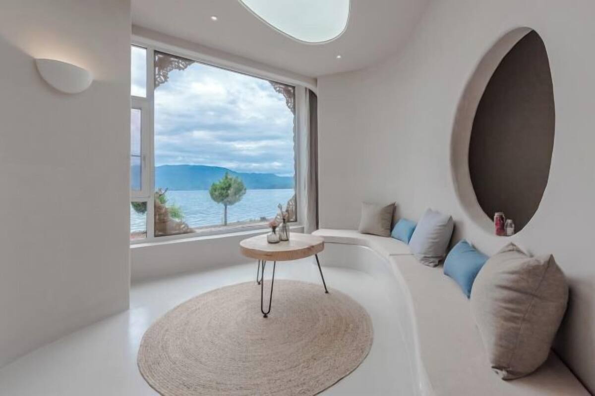 Quarto moderno com uma janela grande, um tapete redondo, um sofá branco com almofadas e um espelho oval fixado na parede.