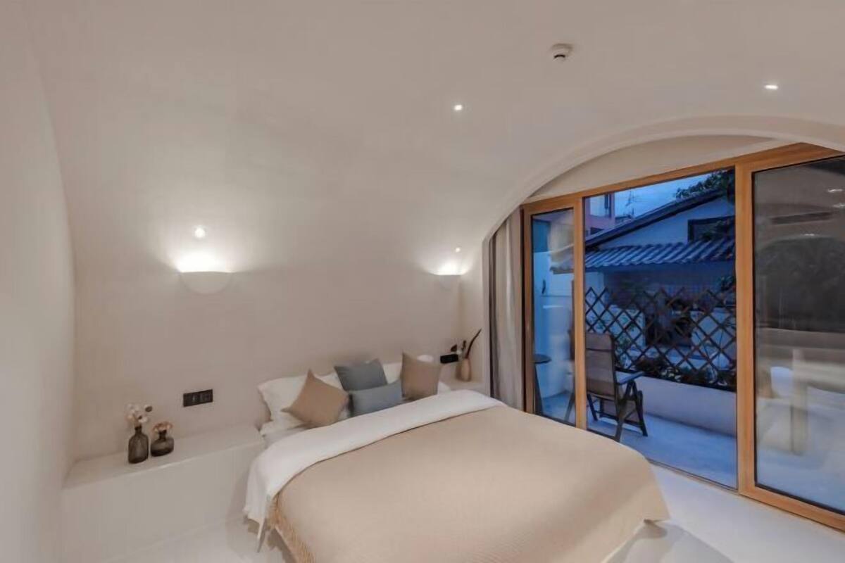 Quarto com cama, mesa de cabeceira e vista para o exterior.