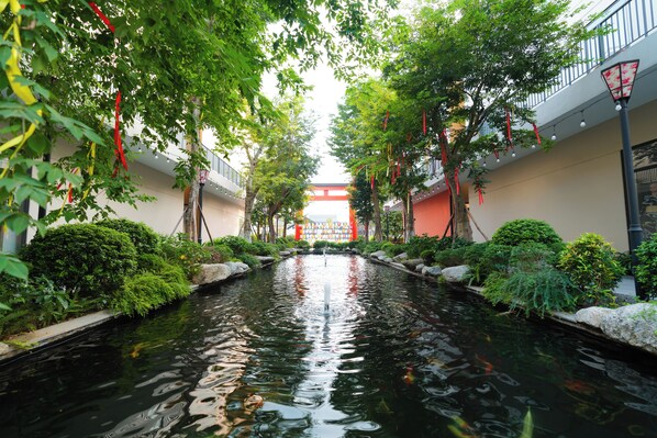 Uma via navegável calma em ambiente interno, ladeada por vegetação exuberante e arquitetura tradicional, que conduz a um torii vermelho.