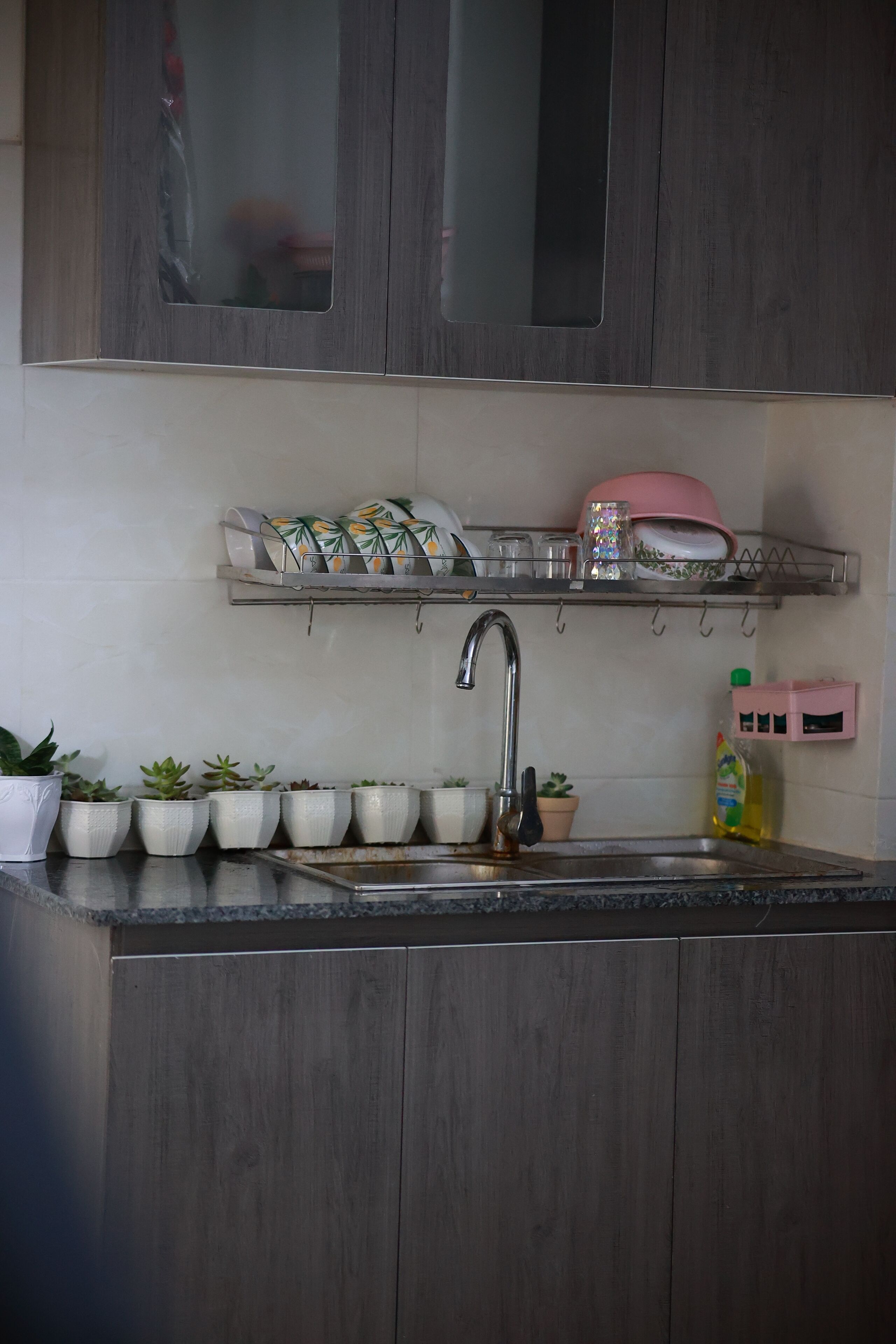 Uma cozinha com uma pia de aço inoxidável, armários de madeira e uma prateleira com louças e uma tigela rosa.
