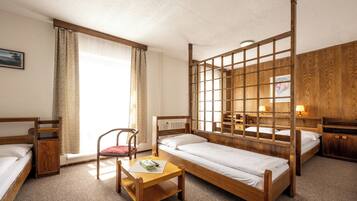 Um quarto com duas camas de madeira, uma escrivaninha de madeira, uma cadeira e uma janela grande com cortinas.