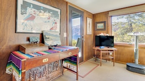 Quarto com escrivaninha de madeira, televisão, pequena mesa com cadeira e uma grande janela com vista para árvores.