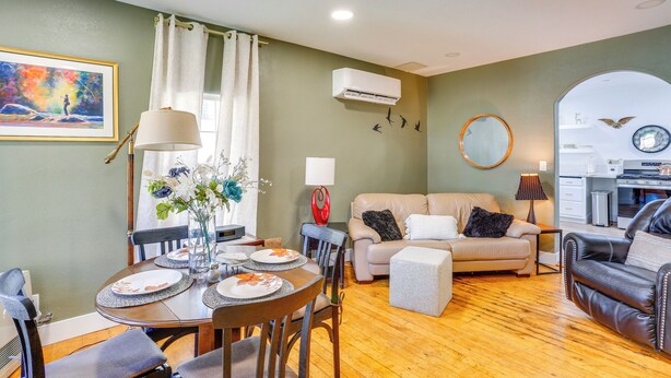 Uma sala de estar com área de jantar, um sofá, uma poltrona de couro, uma cozinha ao fundo e um aparelho de ar condicionado na parede.