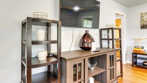 Uma sala de estar com uma mesa consola de madeira escura, uma grande TV de tela plana na parede e uma pequena mesinha de apoio com uma luminária e objetos decorativos.