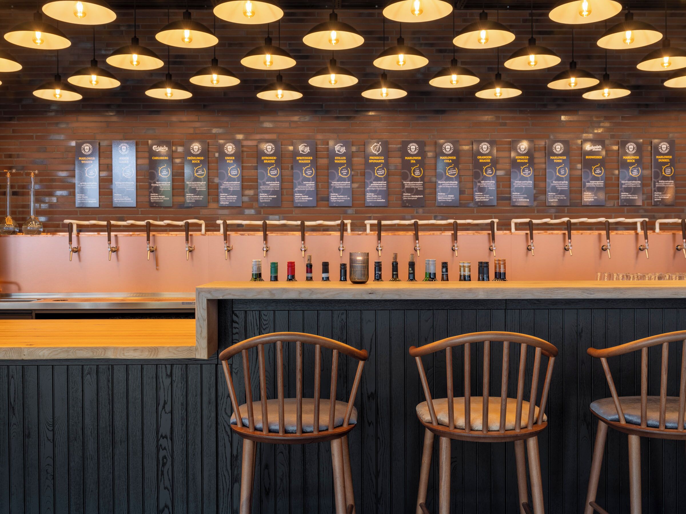 Uma área de bar com várias torneiras de cerveja, cadeiras de madeira e luminárias pendentes.
