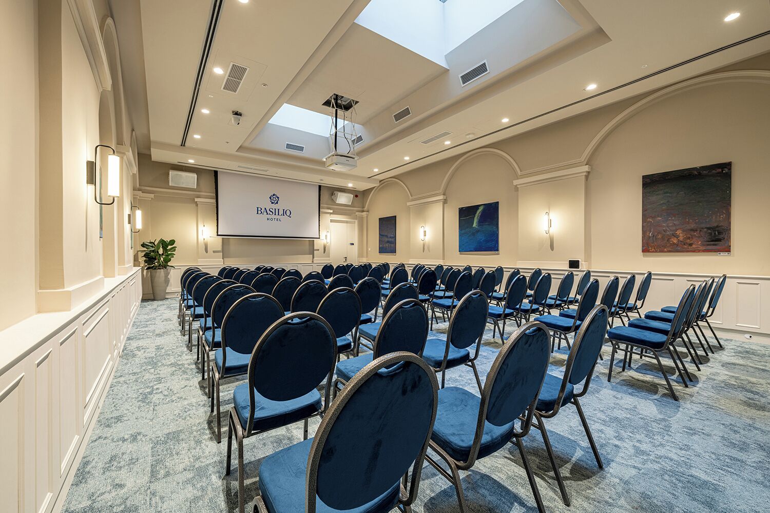 Uma sala de conferências com fileiras de cadeiras azuis, uma tela grande e um letreiro 'BASILIQ HOTEL'.