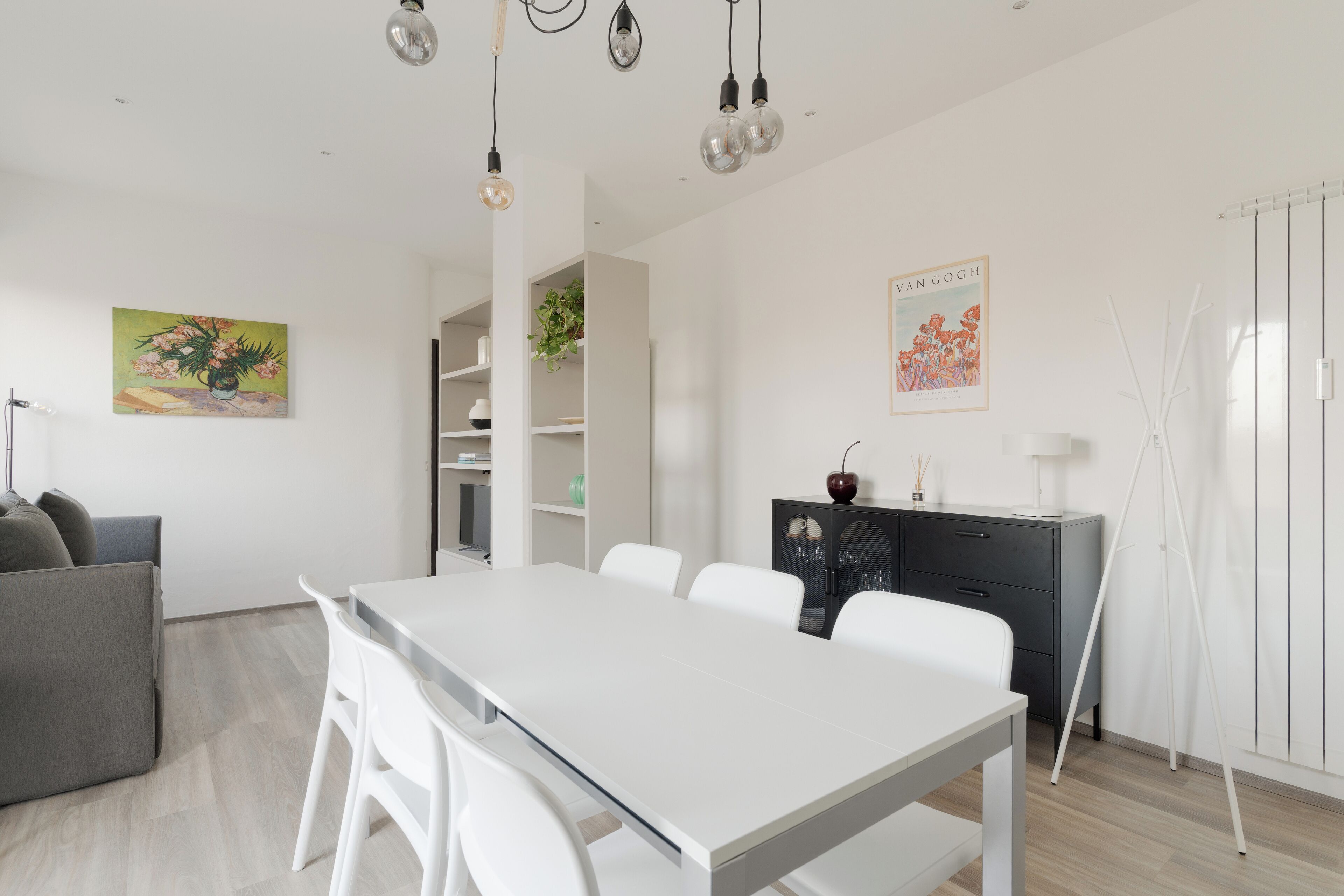 Uma área de refeições moderna com mesa e cadeiras brancas, um sofá cinzento e um armário preto com objetos decorativos.