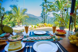 Mesa posta com diversos itens de café da manhã, incluindo torradas, frutas e bebidas, com vista para uma paisagem tropical com palmeiras e montanhas.