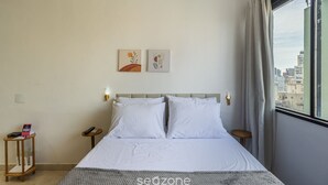 Cama bem arrumada com roupa de cama branca, duas mesas de cabeceira com abajures e uma obra de arte na parede.