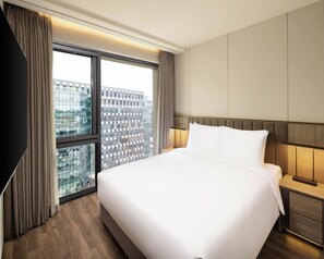 Quarto de hotel com uma cama grande, mesa de cabeceira, televisão e vista para a paisagem urbana através de grandes janelas.
