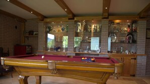 Uma mesa de bilhar em um espaço com paredes de tijolo e vigas de madeira, uma mesa de sinuca e um bar com diversos garrafas e copos.
