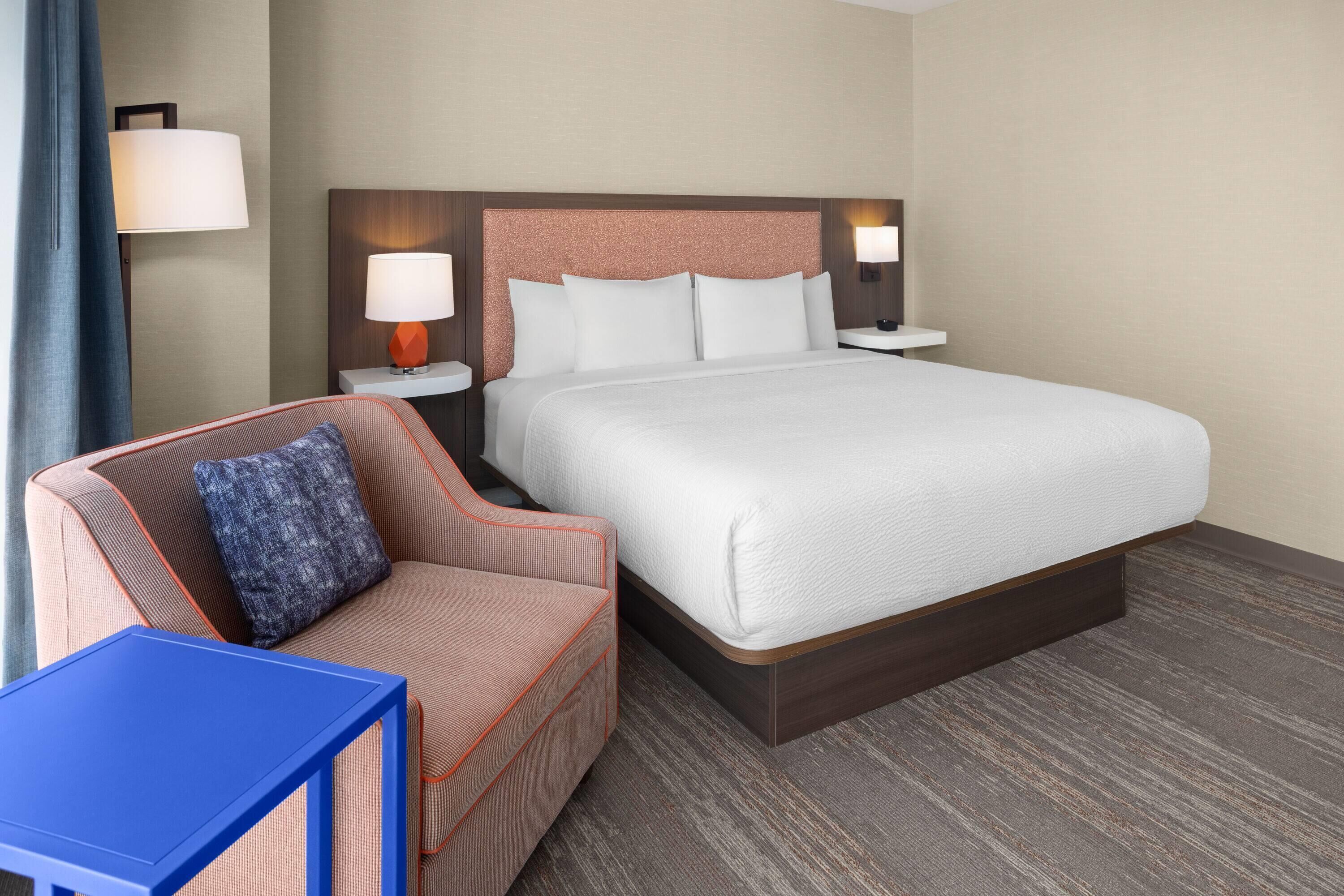 Quarto de hotel com uma cama grande, uma mesa de cabeceira com abajures, uma mesinha lateral azul e uma poltrona marrom com almofada.