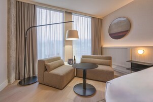Quarto de hotel com duas poltronas, uma mesa redonda pequena, um abajur e uma janela grande com cortinas.