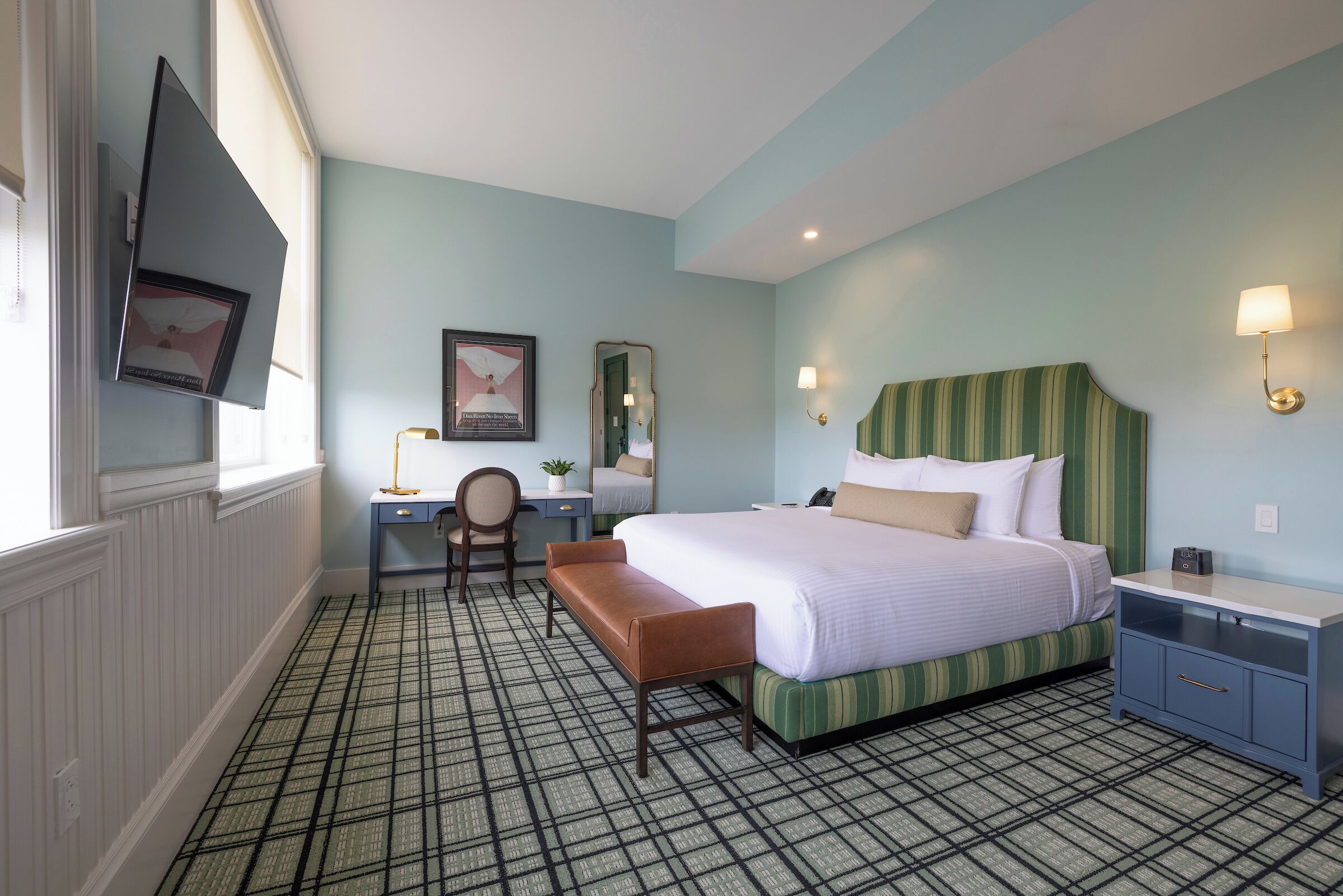 Quarto de hotel com cama, escrivaninha, cadeira, banco, mesa de cabeceira, criado-mudo, TV na parede e espelho.