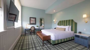 Quarto de hotel com cama, escrivaninha, cadeira, banco, mesa de cabeceira, criado-mudo, TV na parede e espelho.