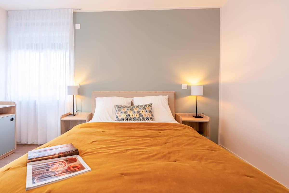Cama bem arrumada com colcha amarela, duas luminárias de cabeceira e um travesseiro com padrão azul e branco.