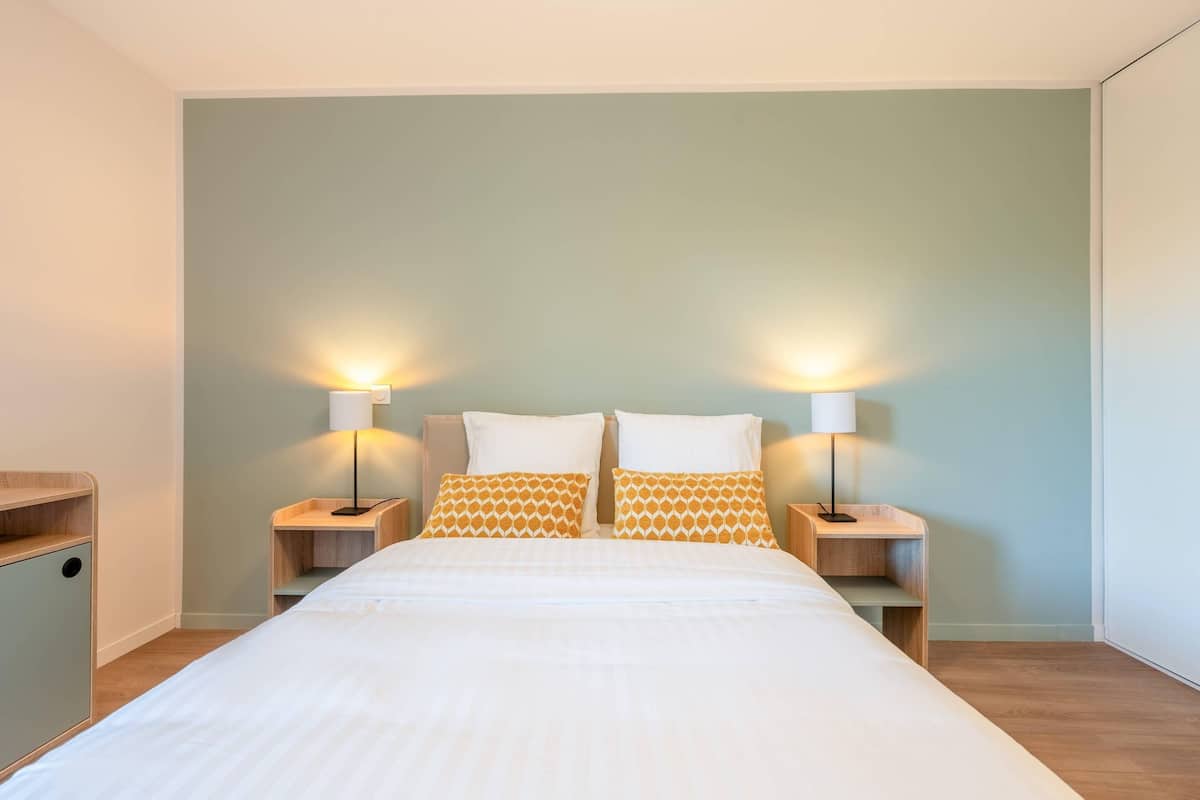 Uma cama bem arrumada com roupa de cama branca e duas almofadas estampadas, acompanhada por duas mesas de cabeceira com abajures, em um quarto com paredes verde-claras e piso de madeira.
