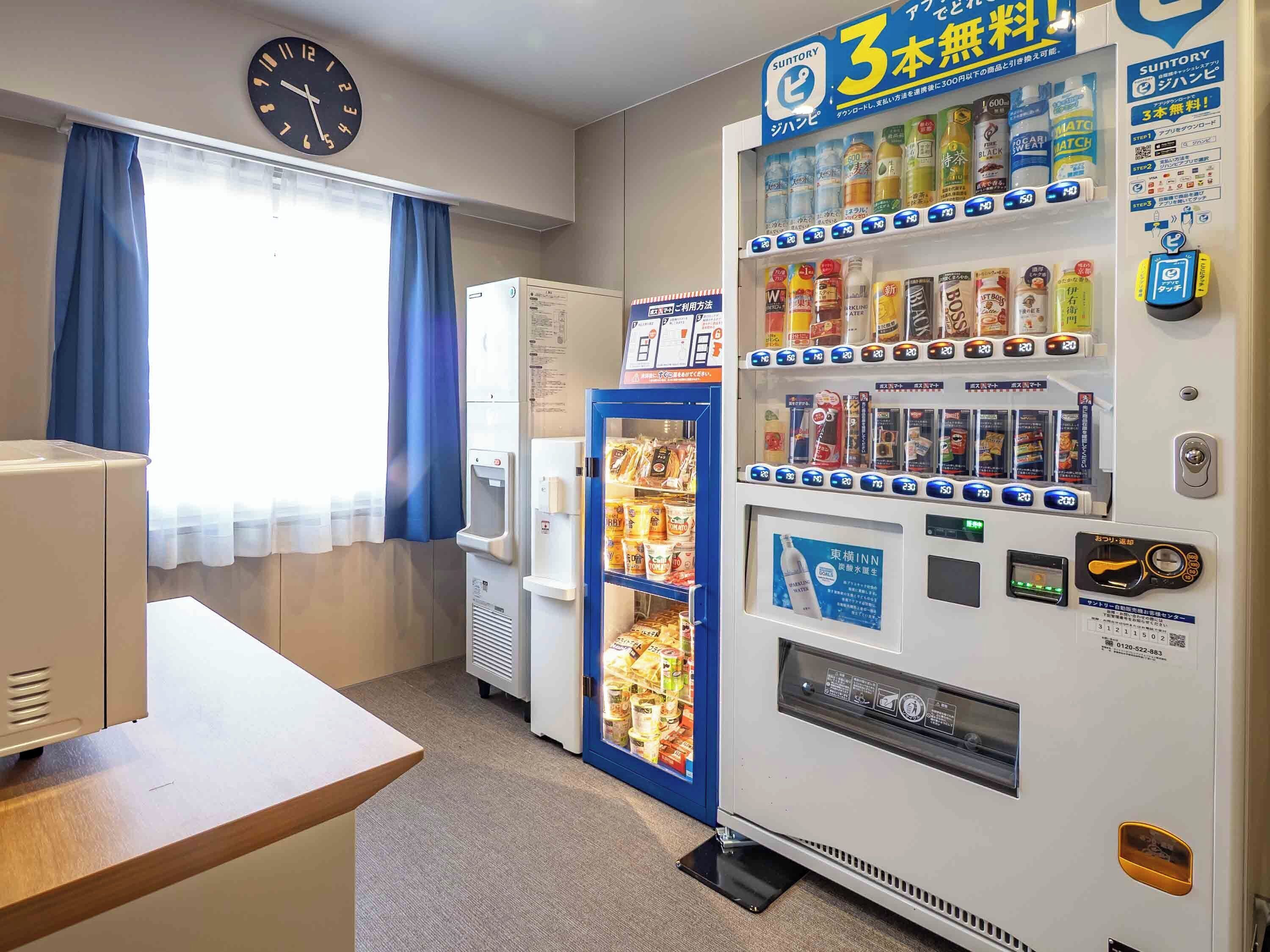 Quarto de hotel com máquina de vendas, mini-geladeira, relógio e janela com cortinas.