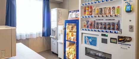 Quarto de hotel com máquina de vendas, mini-geladeira, relógio e janela com cortinas.