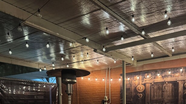 Um interior rústico com uma mesa de madeira redonda, várias cadeiras, paredes de tijolo aparente e luzes em fio.
