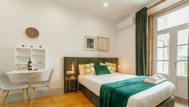 Quarto de hotel com cama, escrivaninha com cadeiras, relógio e janela com cortinas.