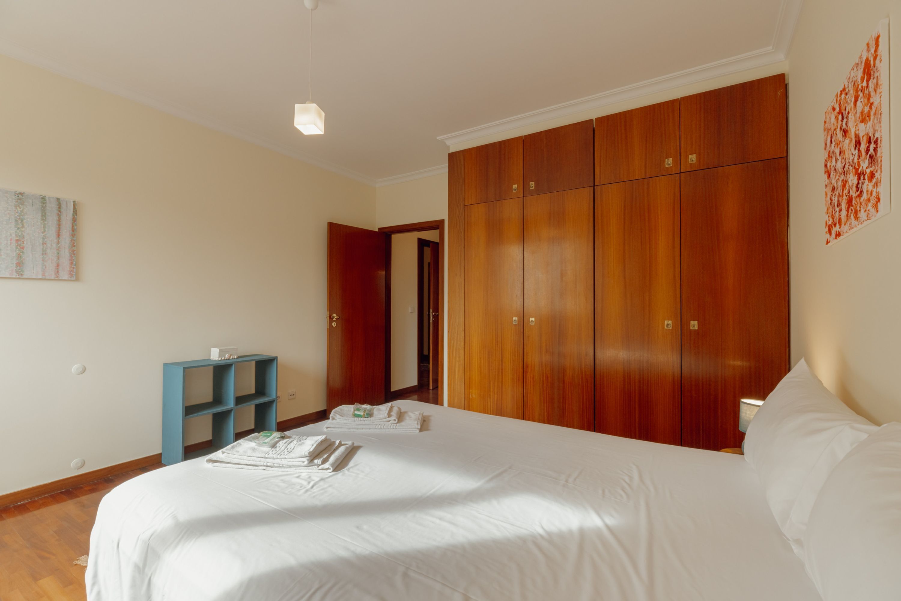 Quarto de hotel com uma cama grande, guarda-roupa de madeira, uma pequena estante azul e uma obra de arte na parede.