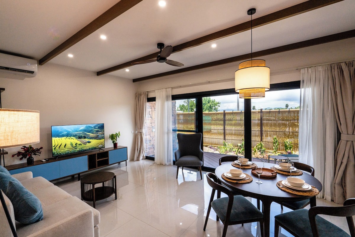 Uma sala de estar moderna com televisão, sofá, mesa de jantar posta para quatro e uma varanda com vista para o campo.