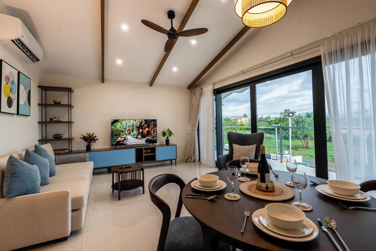 Uma sala de estar moderna com sofá, TV, mesa de jantar posta para quatro e uma grande janela com vista.