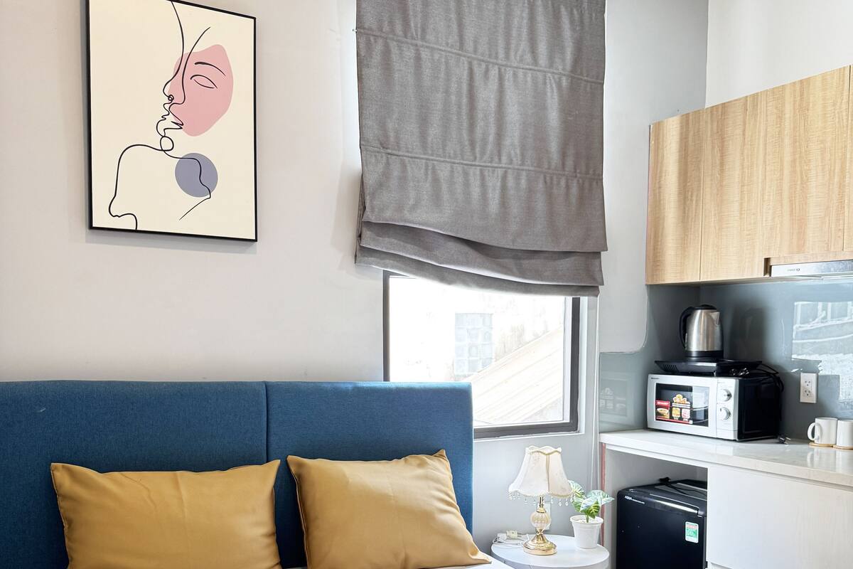 Quarto de hotel com cama, mesa de cabeceira com luminária, pequeno frigorífico, micro-ondas e janela com vista para edifícios.