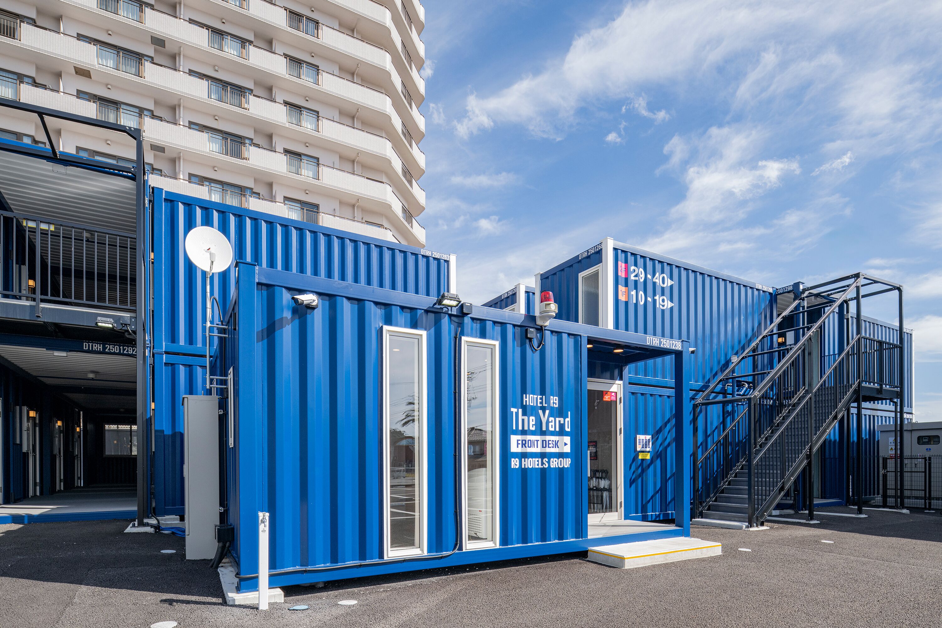 Um hotel container azul chamado "The Yard", com uma receção, escadas e um edifício ao fundo.