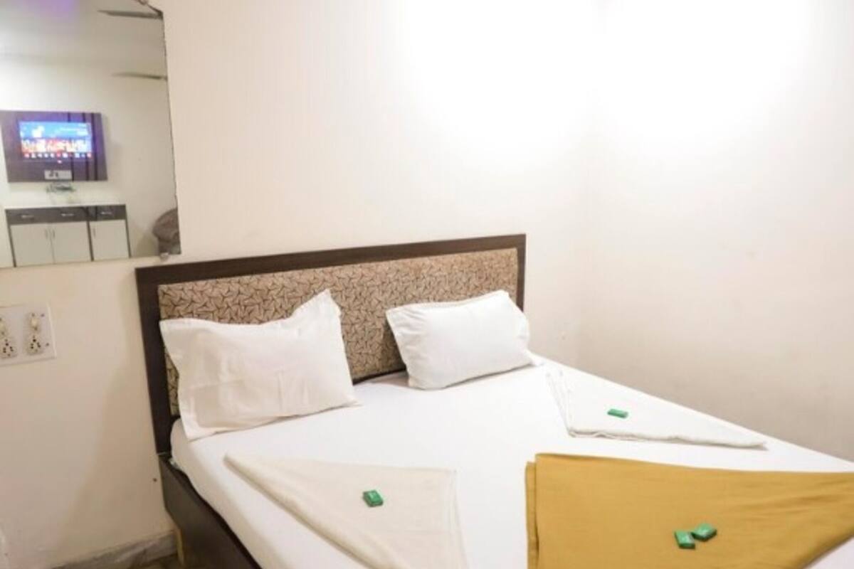 Uma cama bem arrumada com roupa de cama branca e cabeceira marrom, uma pequena TV na parede e uma mesa de cabeceira simples com uma luminária.