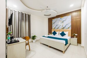 Quarto de hotel com cama, escrivaninha, cadeira, ventoinha no teto e uma grande pintura abstrata na parede.