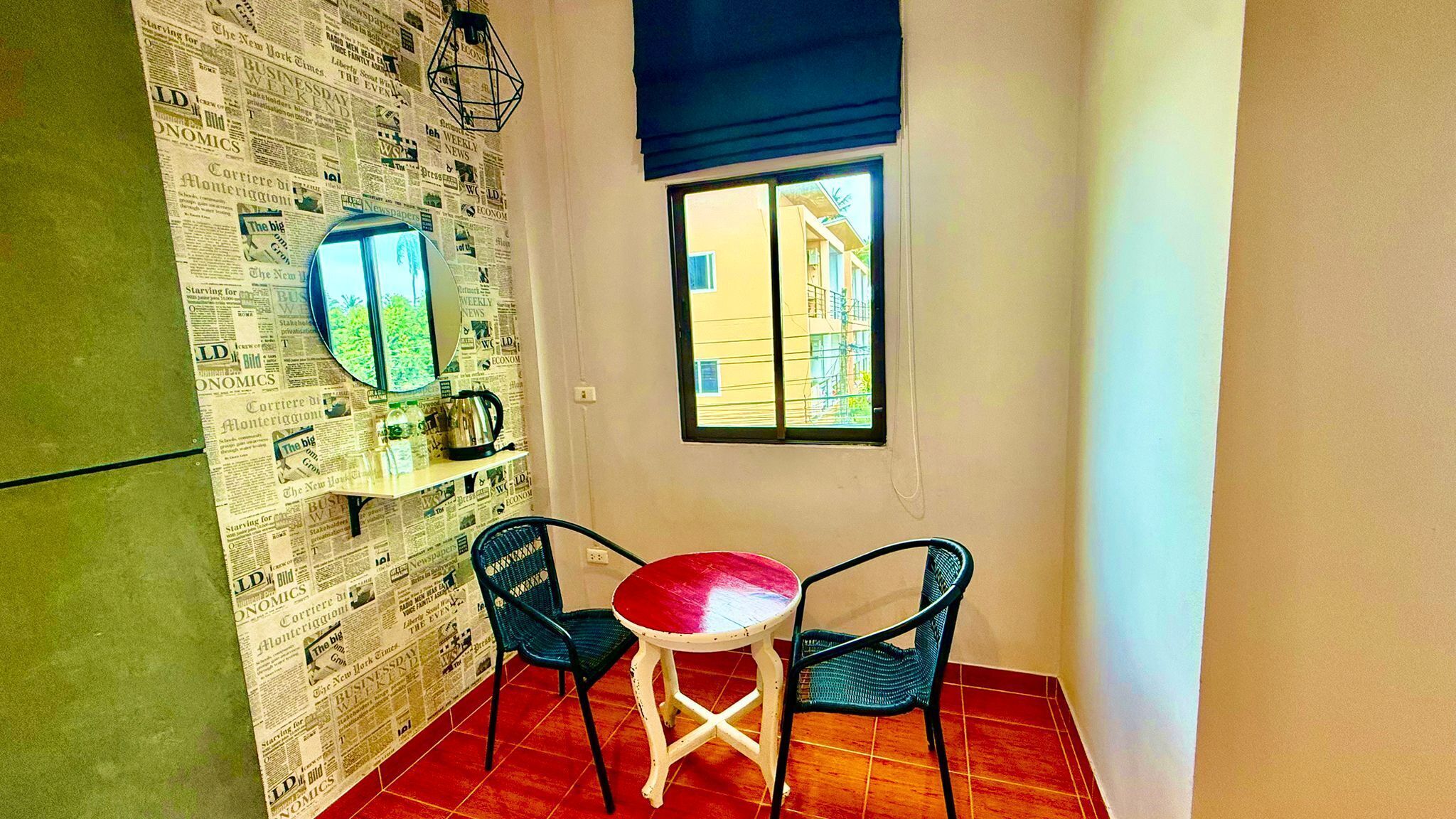 Um quarto pequeno com piso de azulejos vermelhos, uma mesa branca com duas cadeiras, um espelho na parede e uma janela com vista para edifícios.