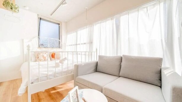 Uma sala de estar iluminada, com um sofá branco, uma mesinha redonda e uma janela com cortinas.