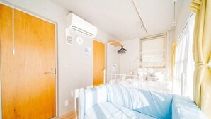 Quarto com porta de madeira, ar condicionado branco, cama branca com roupa de cama azul, radiador branco e janela com persianas.