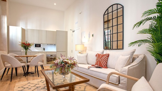 Uma sala de estar moderna com sofá, poltronas, mesa de centro de vidro e cozinha ao fundo.