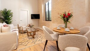 Uma sala de estar com sofá, poltronas, mesa de centro e mesa de jantar com cadeiras. Há uma televisão fixada na parede e uma janela com persianas.