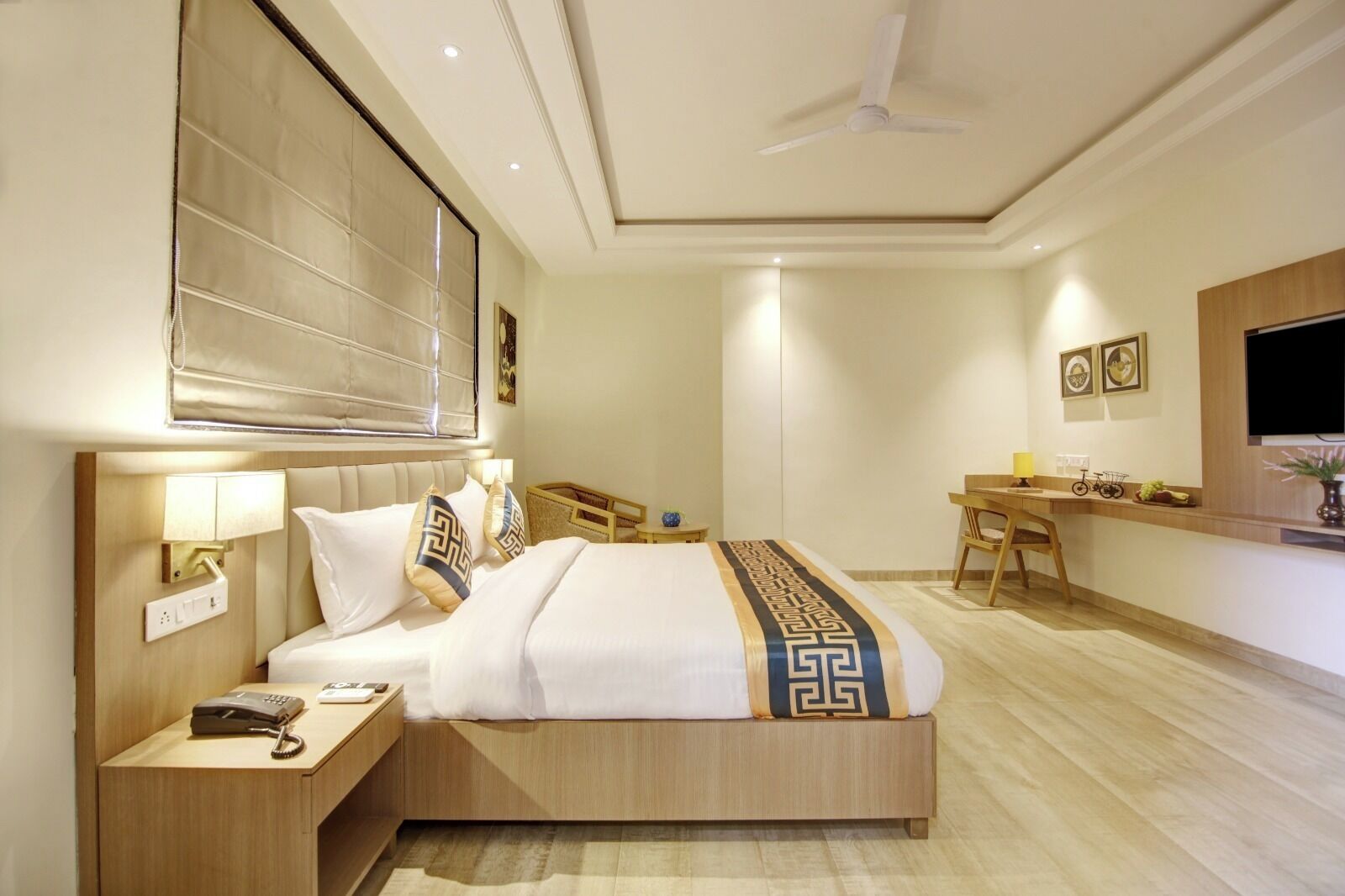 Quarto de hotel com cama, mesa de cabeceira, abajur, escrivaninha, cadeira e televisão na parede.