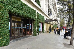 Fachada de um edifício com exterior coberto de folhagem verde, uma grande entrada de vidro e uma passarela para pedestres.