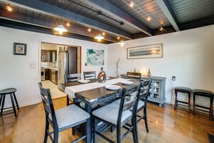 Uma área de refeições com uma mesa de madeira, cadeiras e uma cozinha ao fundo.