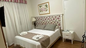 Quarto de hotel com uma cama, mesa de cabeceira com telefone, um criado-mudo e uma cortina.