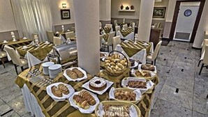 Disposição de buffet com diversos produtos de panificação e doces sobre mesas, organizada em um espaço amplo com piso ladrilhado e decoração neutra.