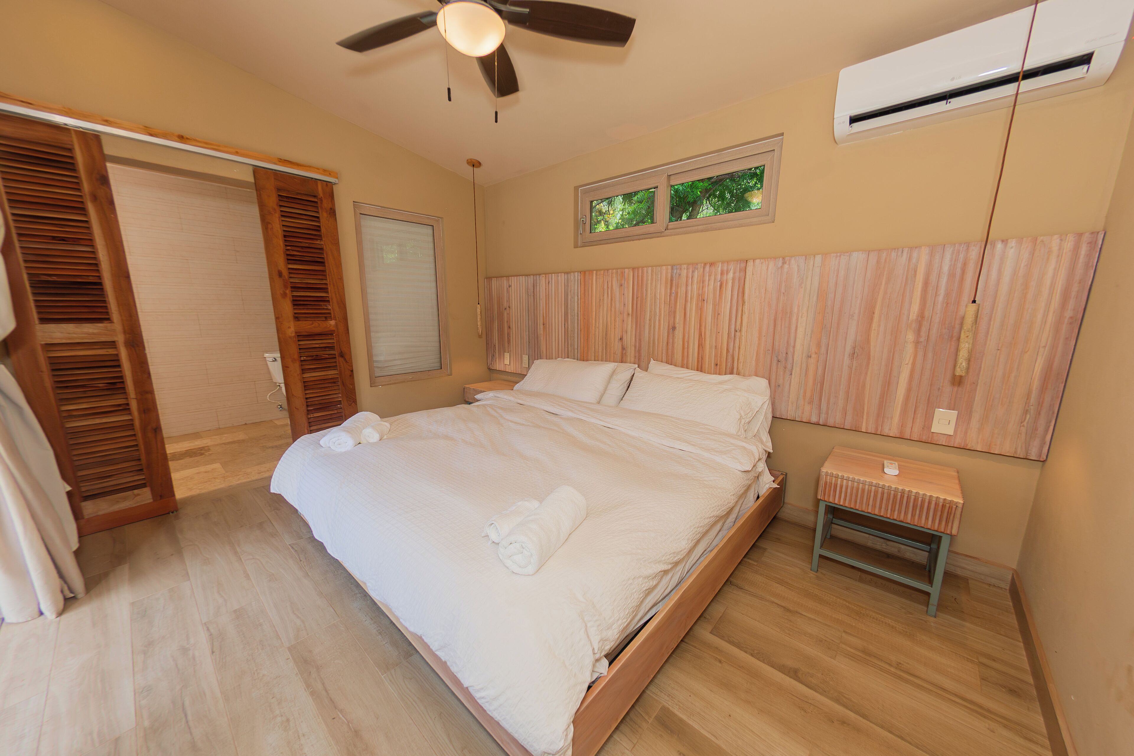Quarto com cama, piso de madeira, ventilador de teto, ar-condicionado e janela com vista para o verde.