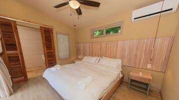 Quarto com cama, piso de madeira, ventilador de teto, ar-condicionado e janela com vista para o verde.