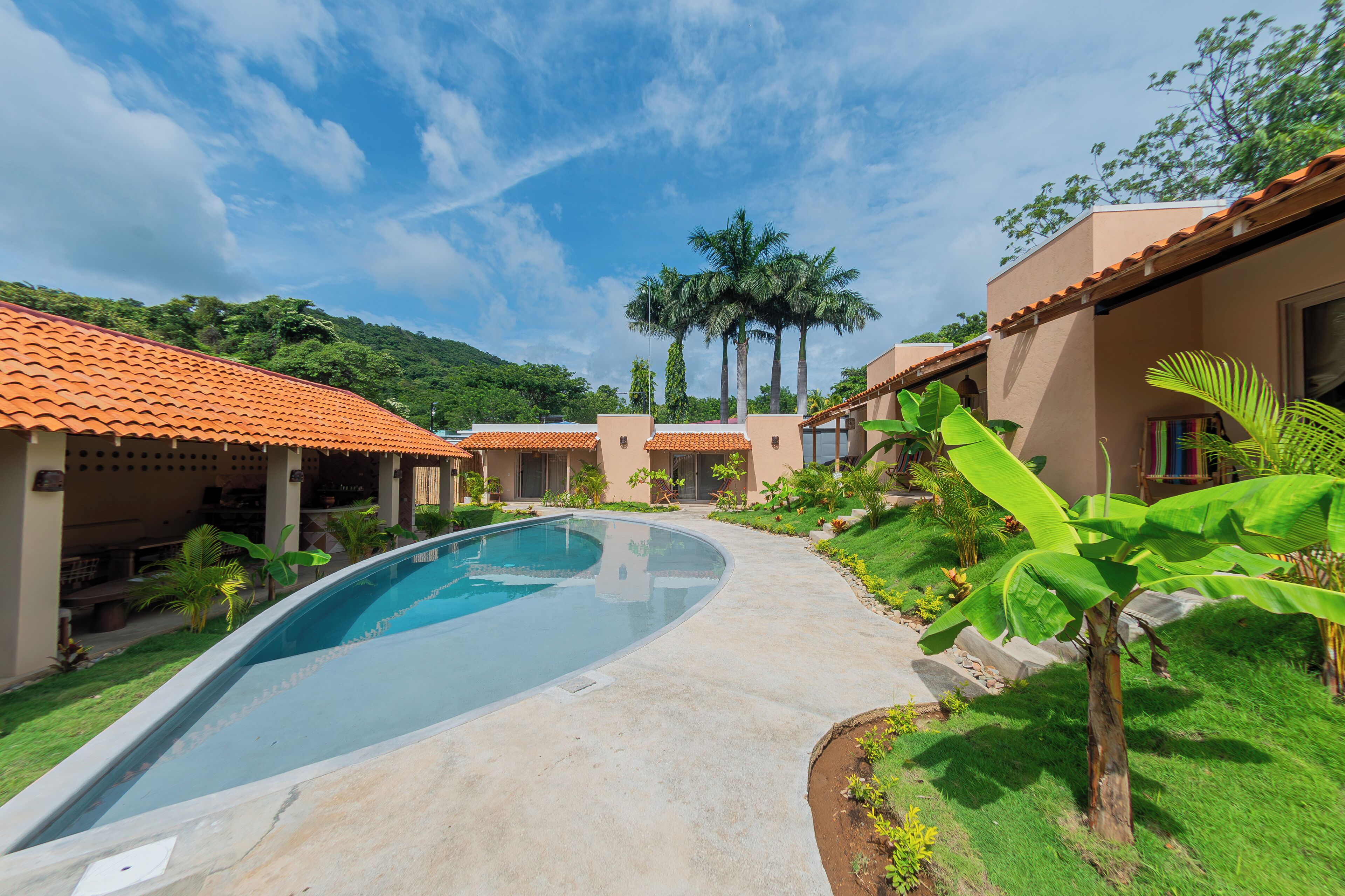 Uma área de piscina com borda curva, rodeada por vegetação e palmeiras, com edifícios com telhados vermelhos ao fundo.