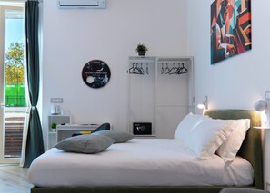 Quarto com cama, almofadas, ar condicionado fixado na parede, relógio de parede, uma pequena planta, mesa de cabeceira com abajur e janela com cortinas.