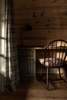 Interior de uma cabana de madeira com cortina xadrez, uma escrivaninha com abajur e uma cadeira de madeira.