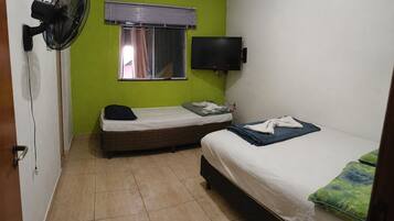 Quarto de hotel pequeno e individual, com uma cama, televisão, janela com persianas, ventilador e porta.