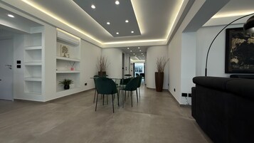 Uma sala de estar moderna com um sofá preto, uma mesa de jantar de vidro com cadeiras verdes e uma grande planta em vaso.